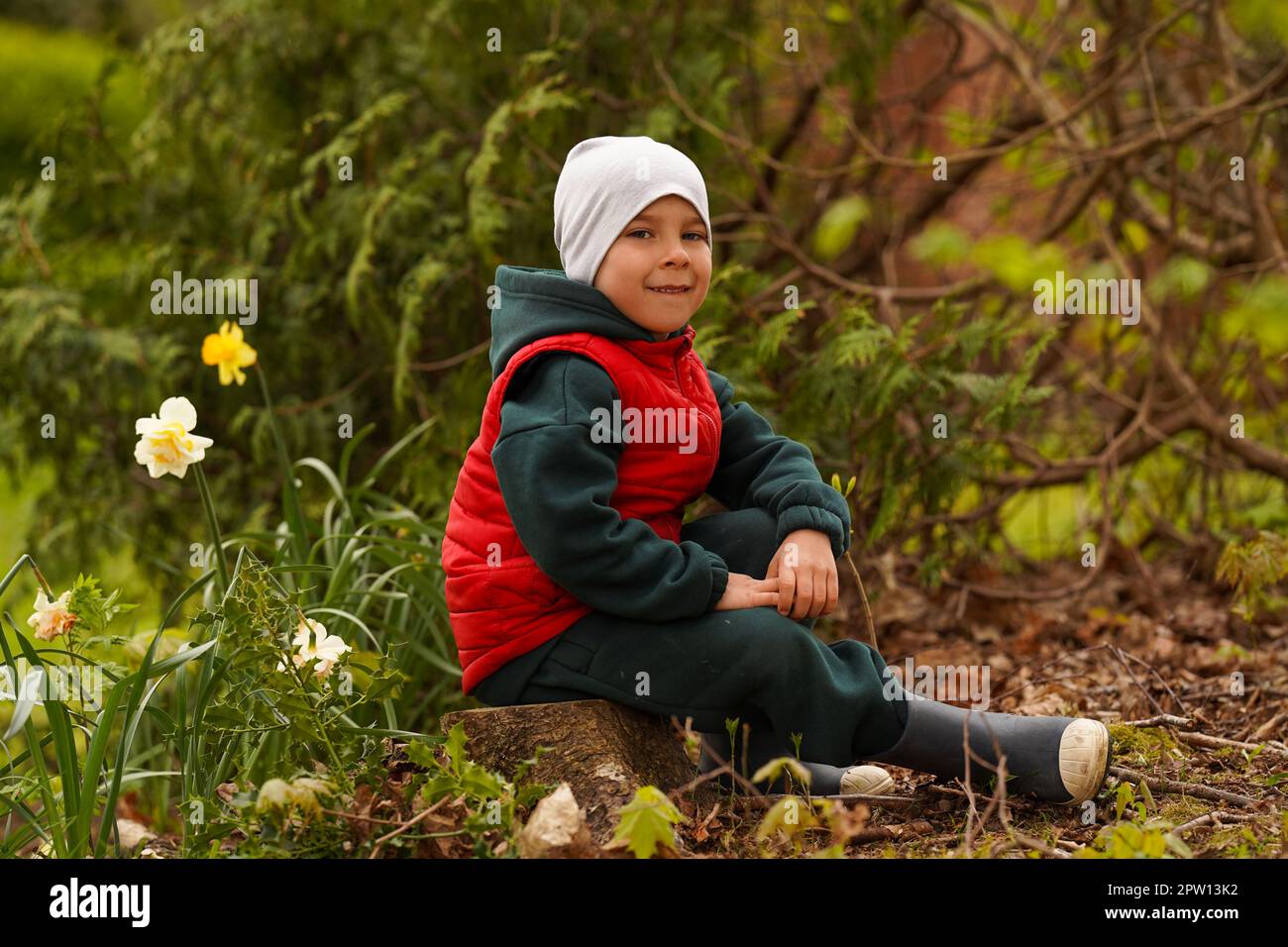A little boy in spring clothes sits on a tree stump among flowers and ...
