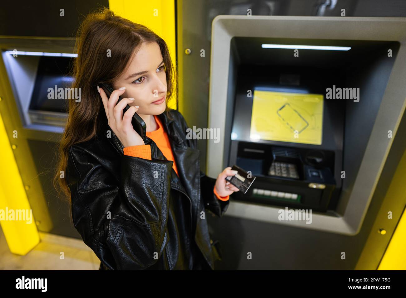 Young brunette girl withdrawing money from credit card at yellow ATM ...