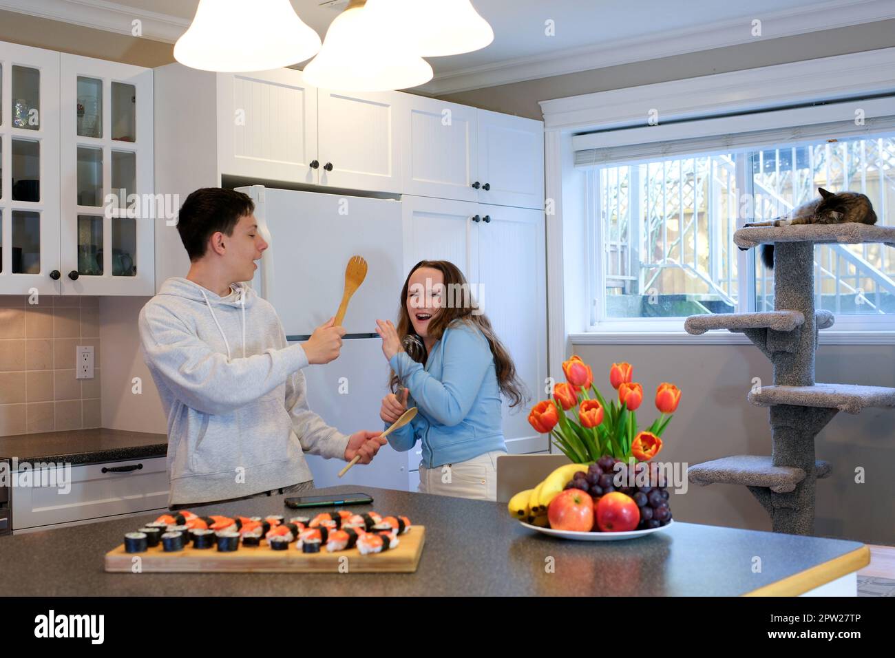 Protect yourself. Excited boy and girl foolishly imitate a battle with kitchen utensils standing in modern kitchen. Funny teenagers are fooling around playing with a wooden spatula and whisk Stock Pho...