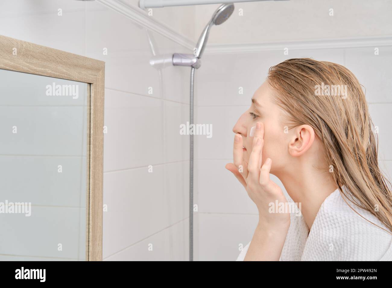 Young woman standing in profile and applying moisturizing cream on face ...