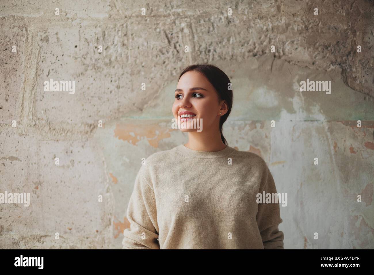 Happy young woman in beige jumper smiling and looking away while ...