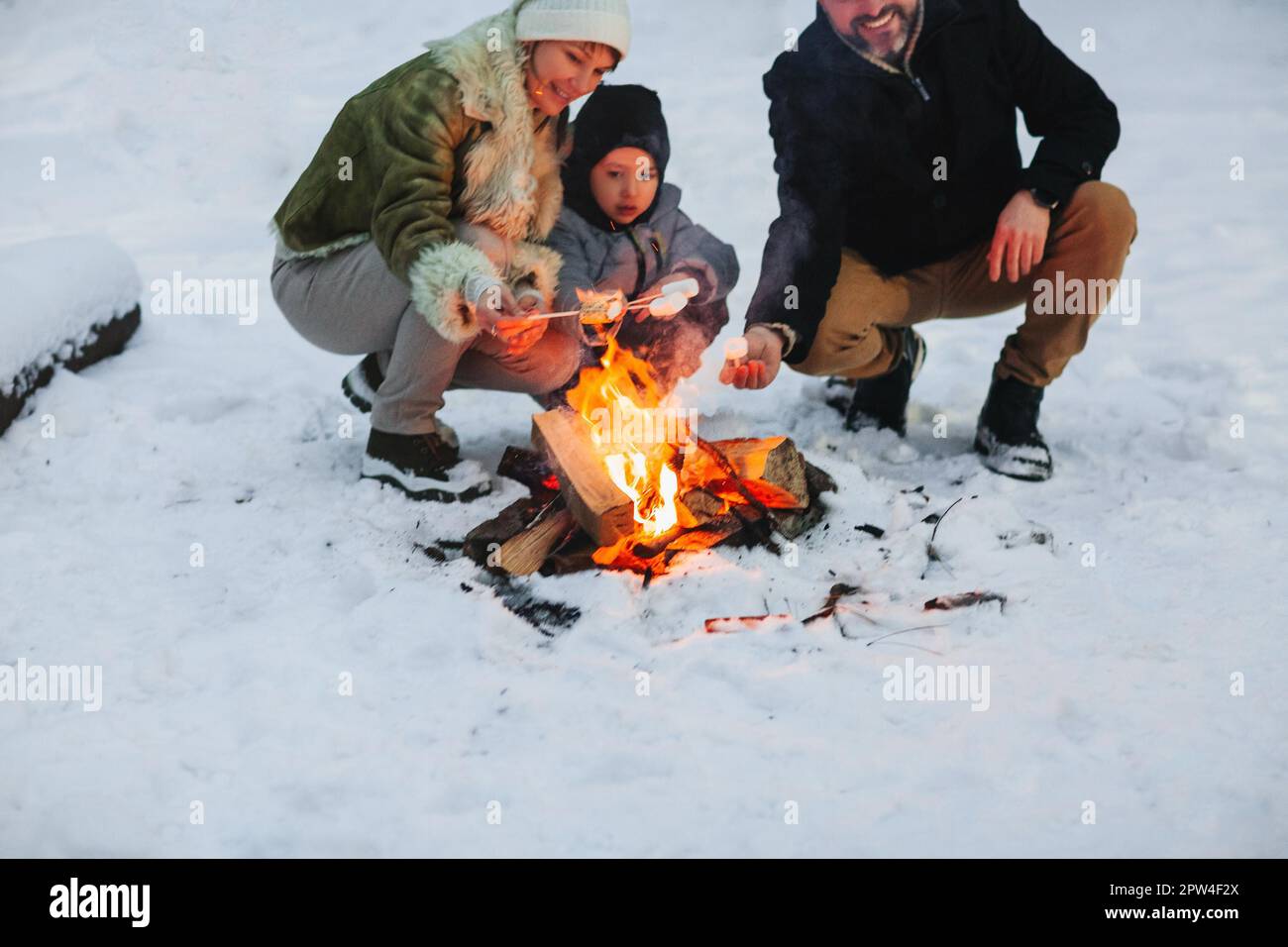 Cheerful parents and son enjoying hot drink while resting near campfire in evening in winter ...