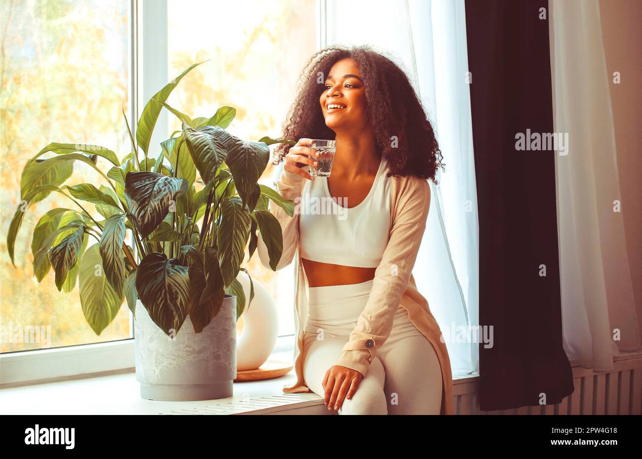 Young positive african american woman sitting on windowsill with glass of pure mineral water and ...