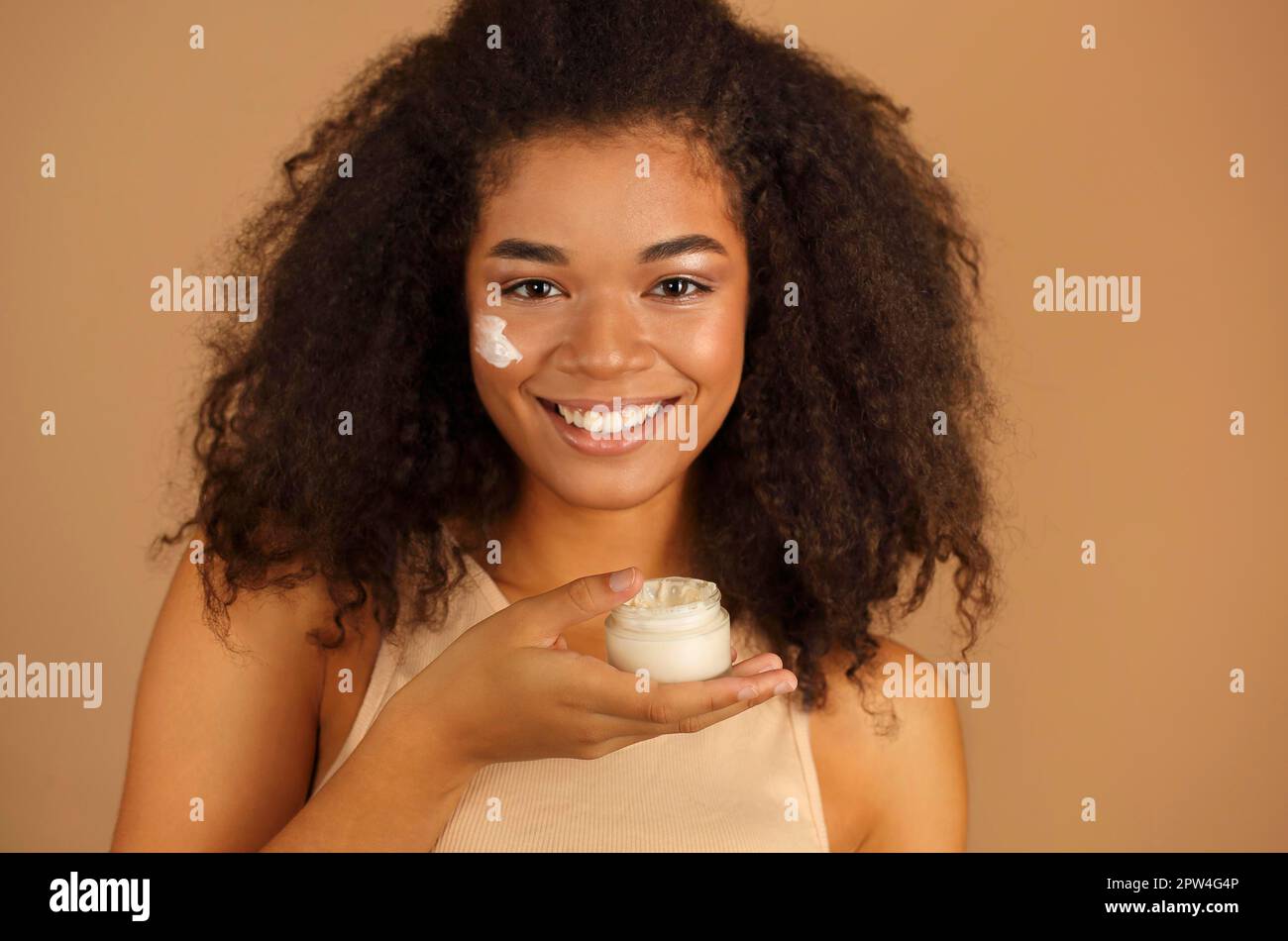 Smiling dark skinned woman with curly afro hair applies face cream on ...