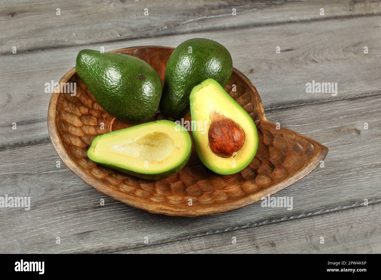 Three avocados, one of them cut to half, seed visible, in carved wooden ...
