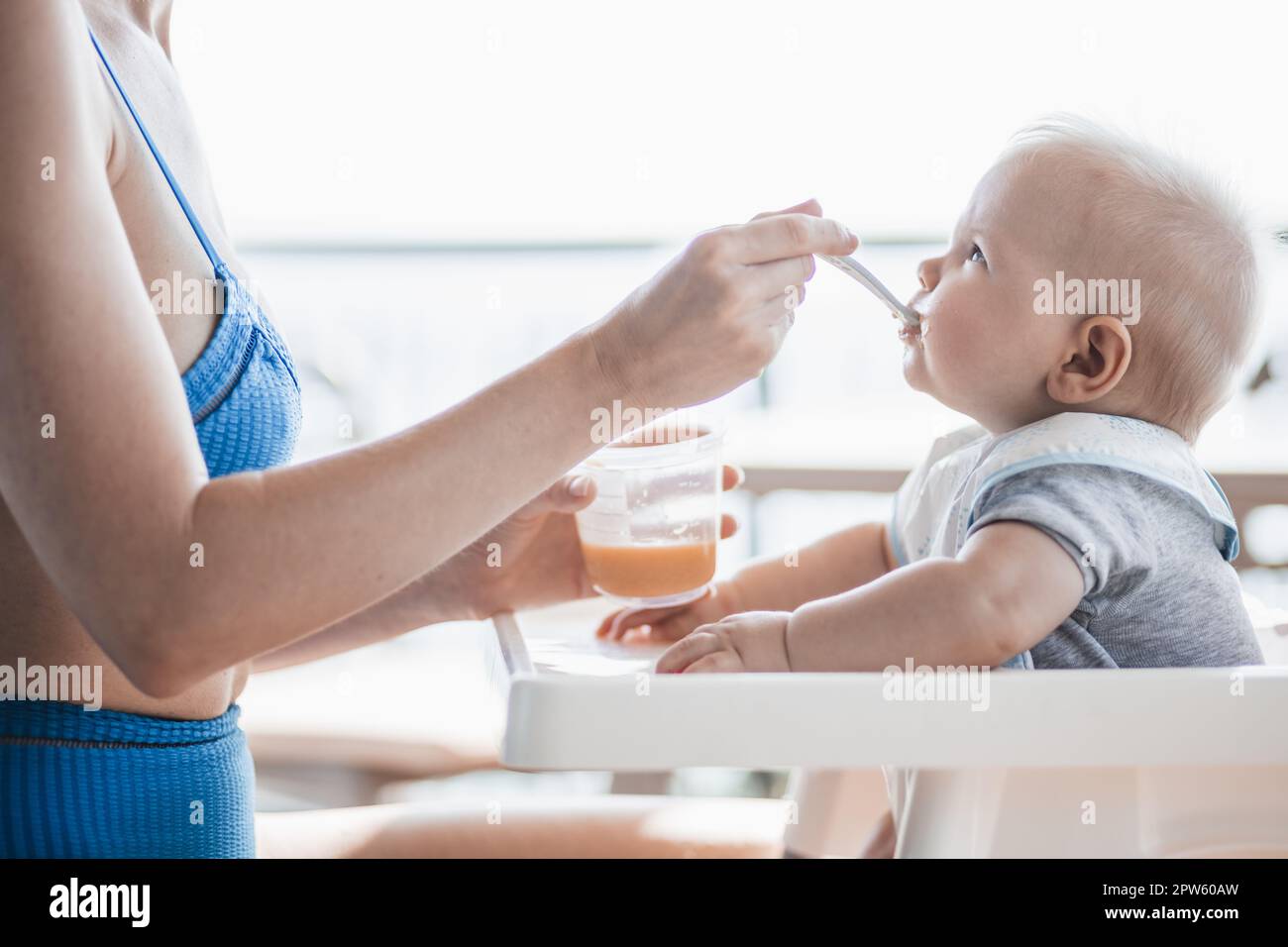 Mother spoon feeding her baby boy child in baby chair with fruit puree ...