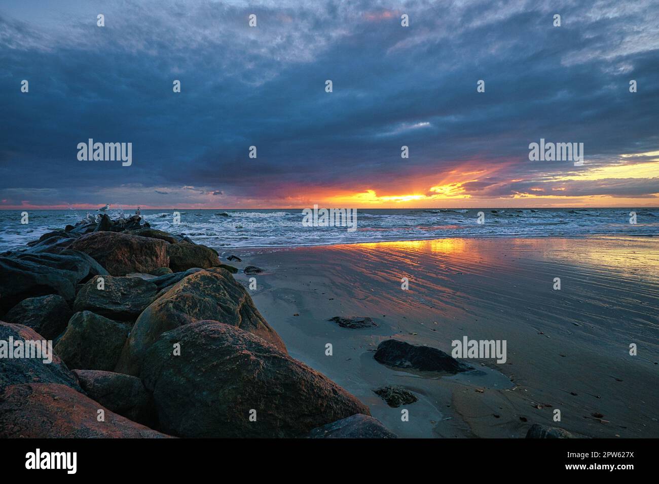 Sandy beach with stone dunes reaching into the sea. Dramatic sunset. On ...