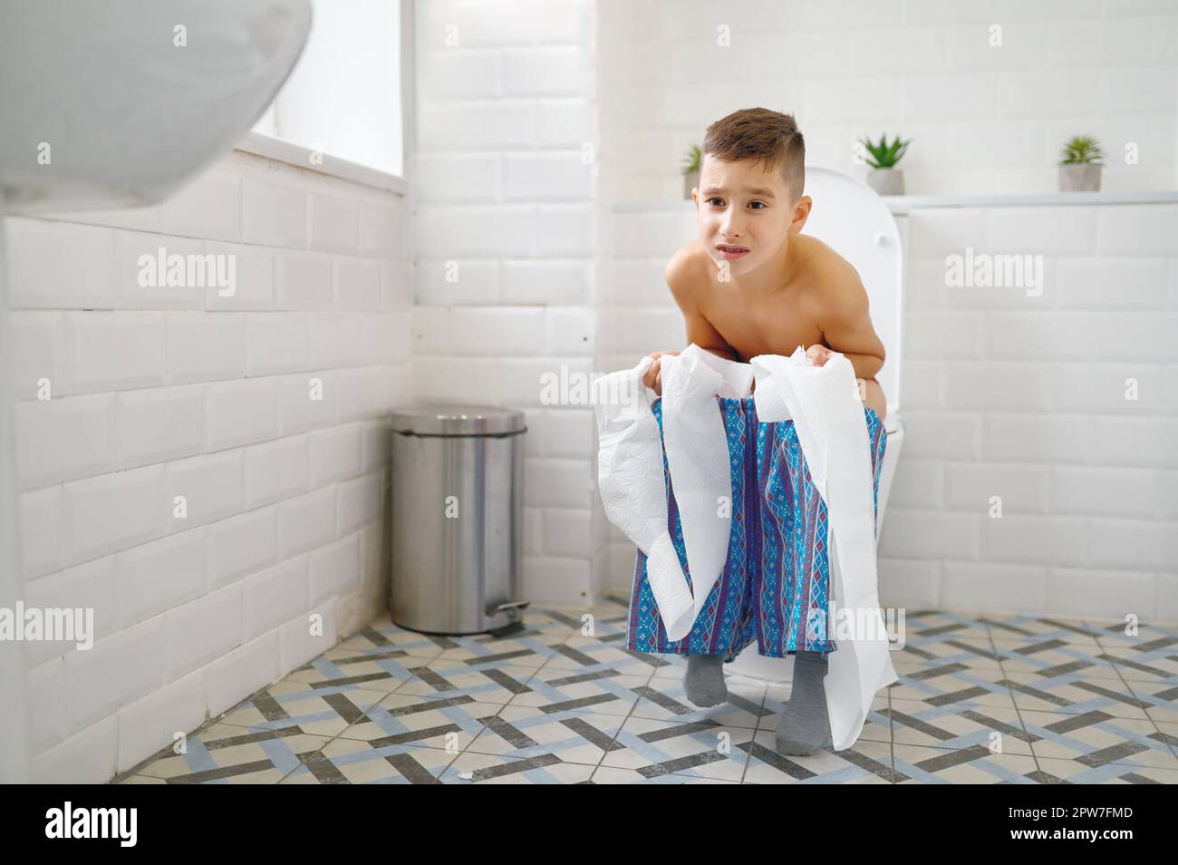 Frowning little boy holding paper sitting on toilet and trying to poop ...
