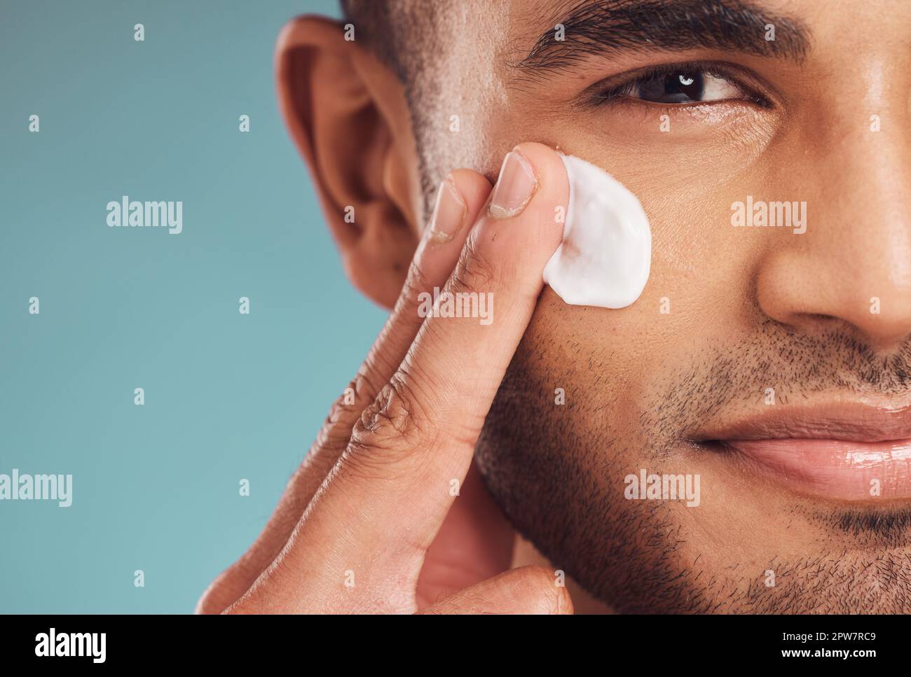Closeup of one young indian man applying moisturiser lotion to his face while grooming against a ...