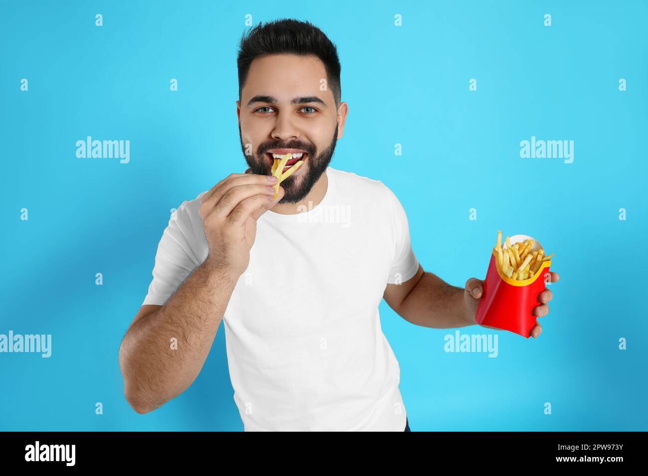 Young man eating French fries on light blue background Stock Photo - Alamy