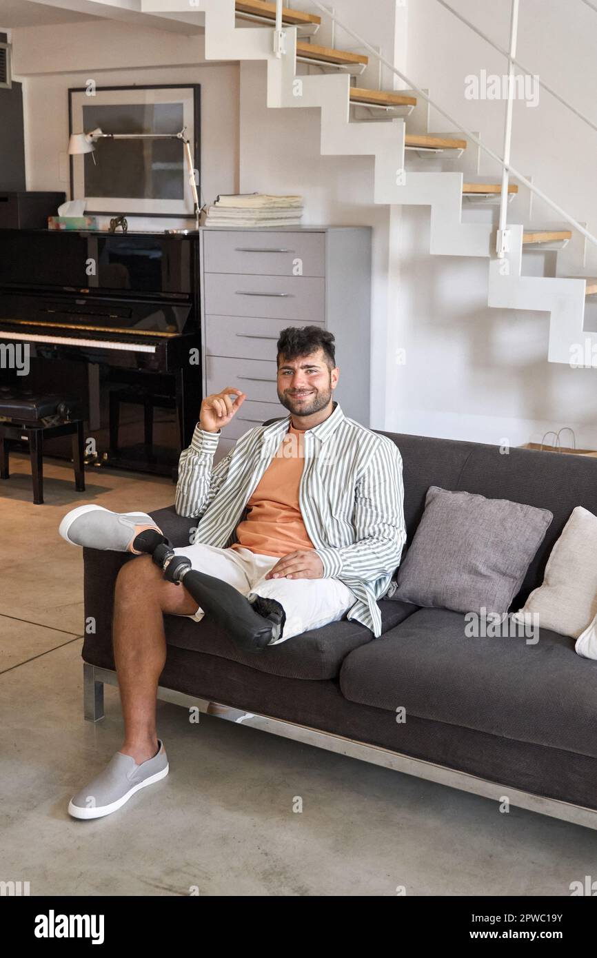 Happy young man with above knee leg prosthesis sitting on sofa at home ...