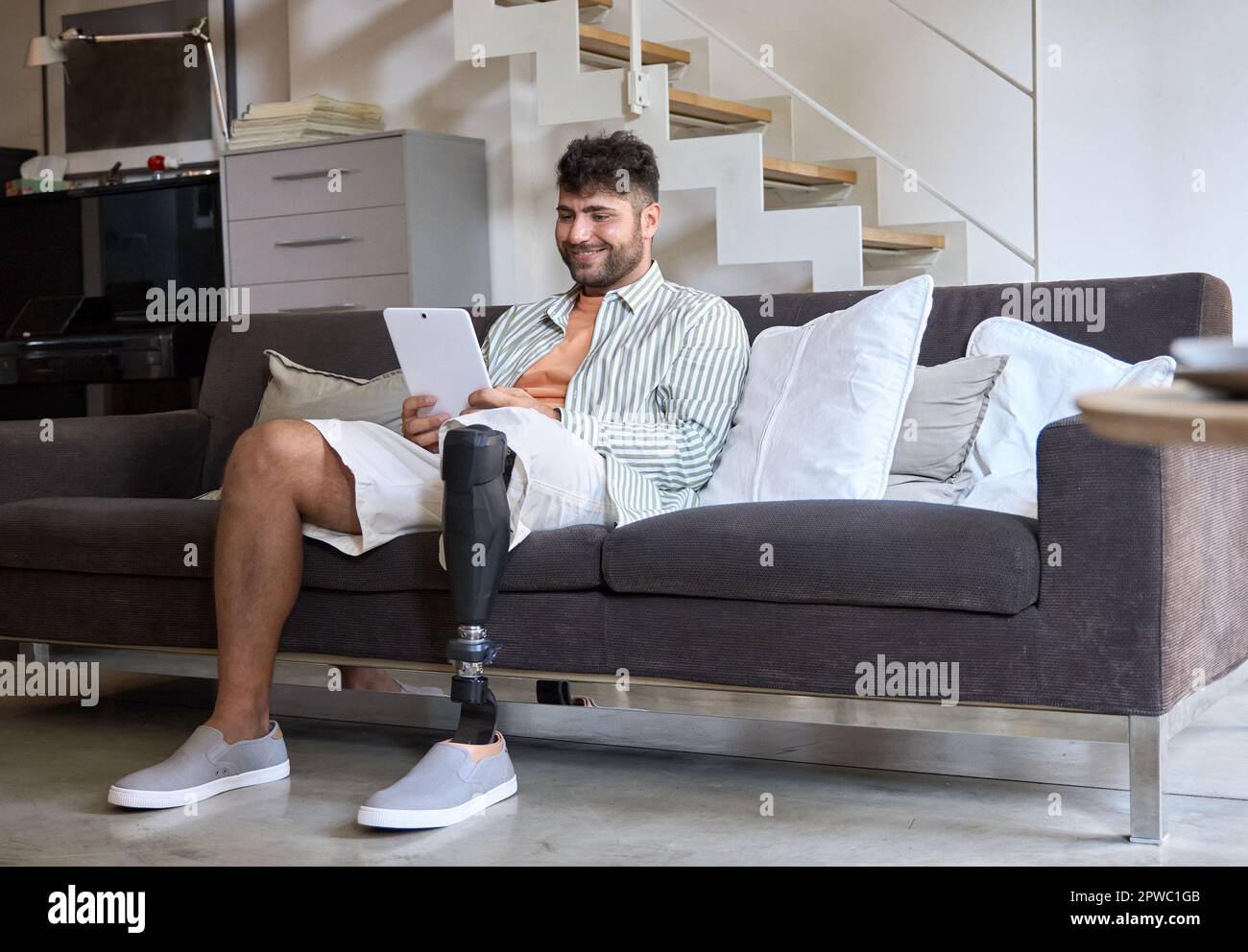 Happy man with disability leg prosthesis sitting on sofa at home using ...