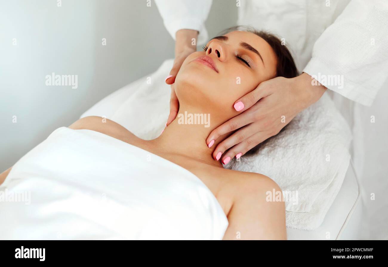 Unrecognizable masseuse in gown standing in spa salon and massaging face of female customer ...