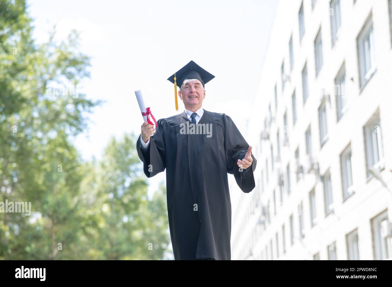 An old man in a graduation gown walks outdoors and holds a diploma ...