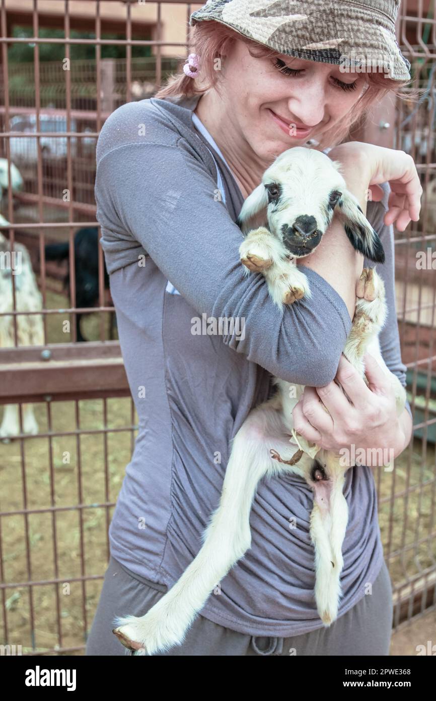 In a farm, a woman sits on a porch with a small baby goat with its tail ...