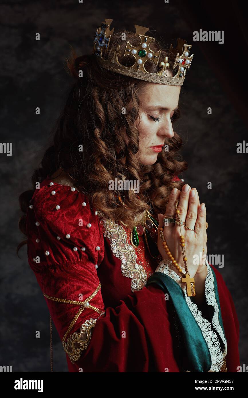 medieval queen in red dress with rosary and crown praying on dark gray ...