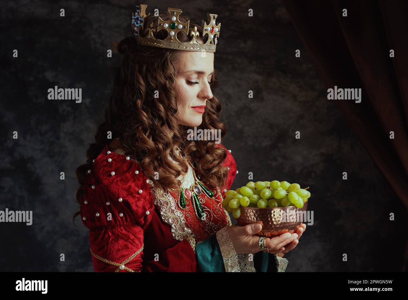 medieval queen in red dress with plate of grapes and crown on dark gray ...