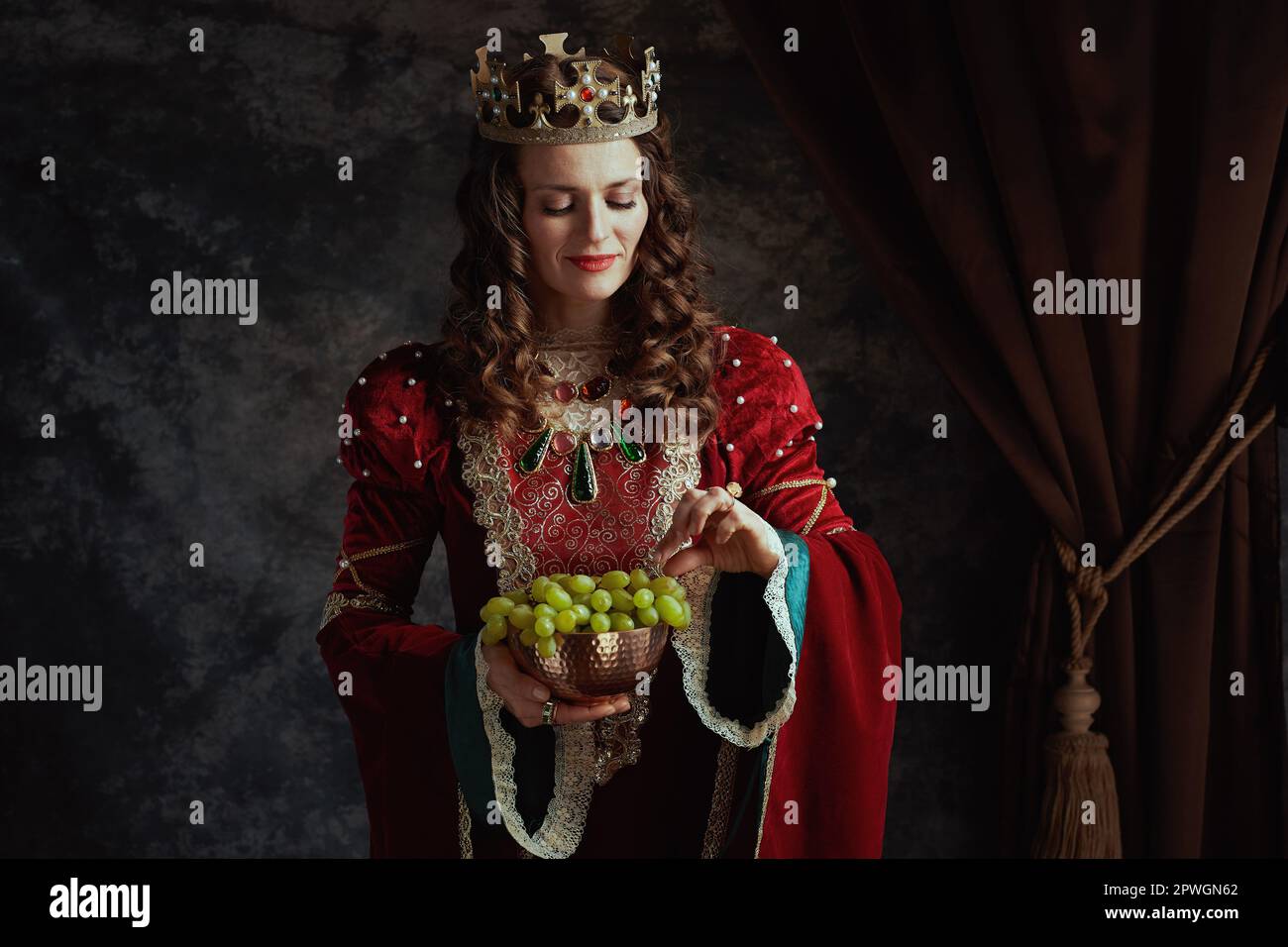 happy medieval queen in red dress with plate of grapes and crown on ...