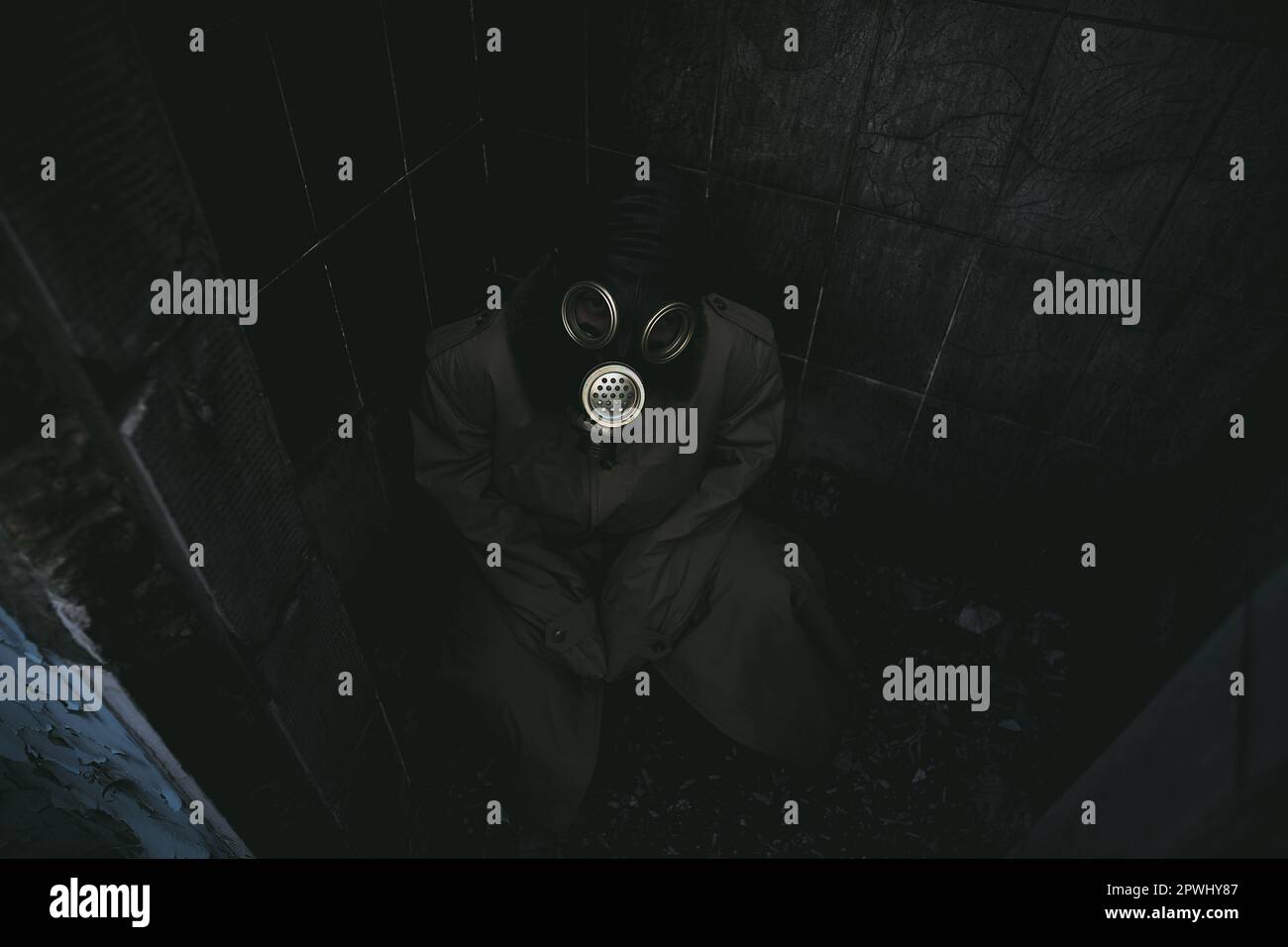 A man in a gas mask sits in a small dark room on his haunches and looks ...