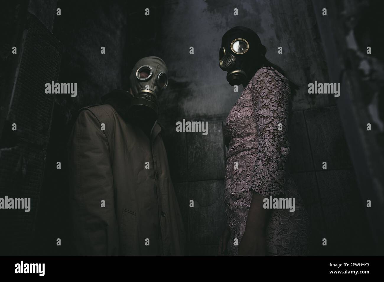 two people in gas masks stand against a wall in a small dark room, low ...