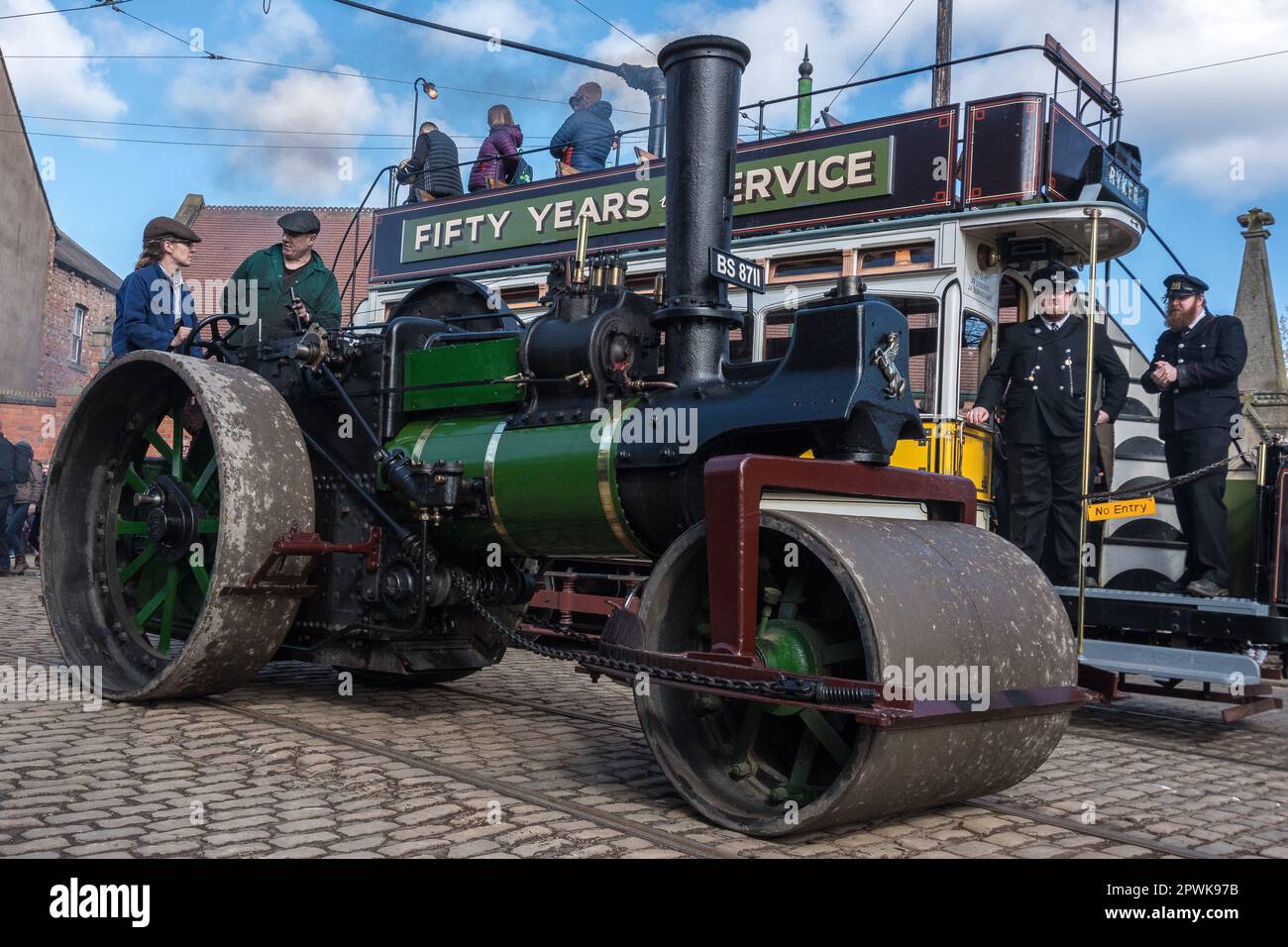 An old steam road roller passing a double decker tram car on a cobbled ...