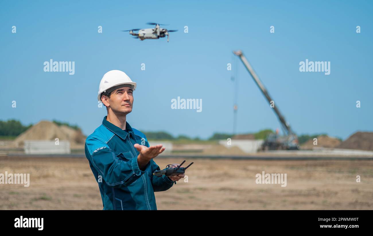 A man in a helmet and overalls controls a drone at a construction site ...