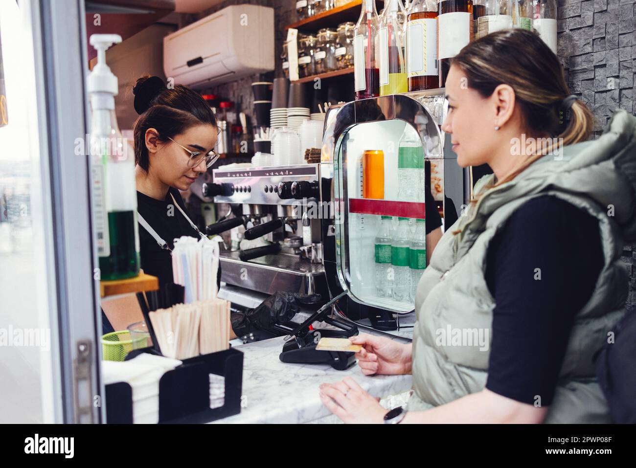 Smiling young barista girl in casual clothes, apron and black gloves ...