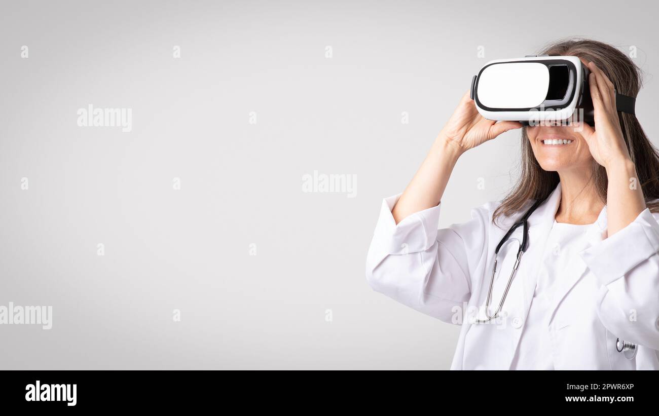 Senior doctor woman wearing virtual reality glasses, standing on grey ...