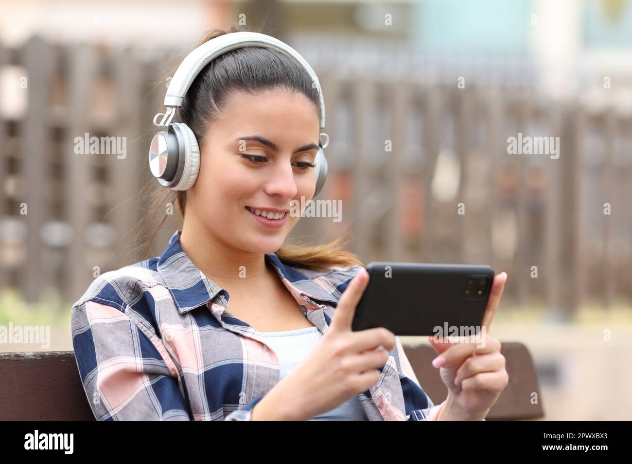 Happy teen wearing headphones watching media on phone in a bench Stock ...