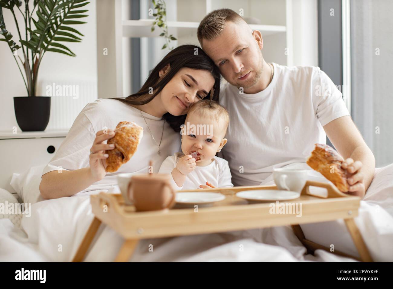 Loving spouses in everyday wear holding croissants while cuddling little daughter that eating ...