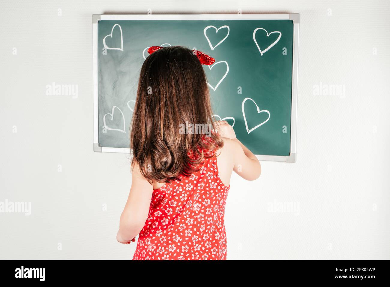 Little girl in red dress turning away draw with chalk hearts on green ...