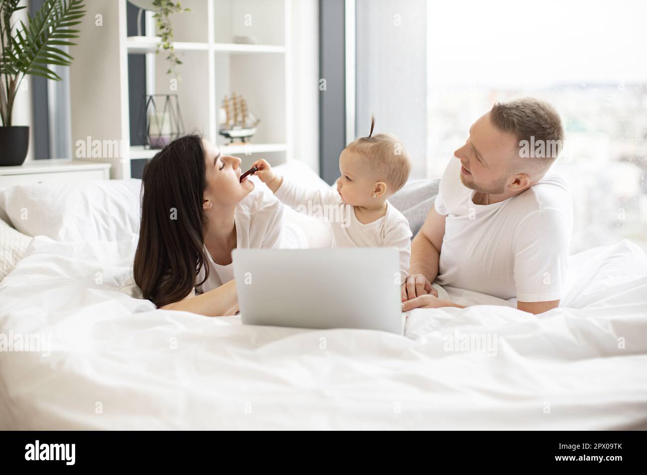 Pretty baby girl on bed feeding cheerful mother with fruit roll while ...