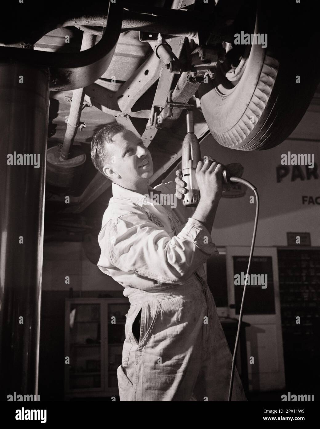 1950s MAN MECHANIC USING AN ELECTRIC WRENCH SERVICING THE UNDER ...