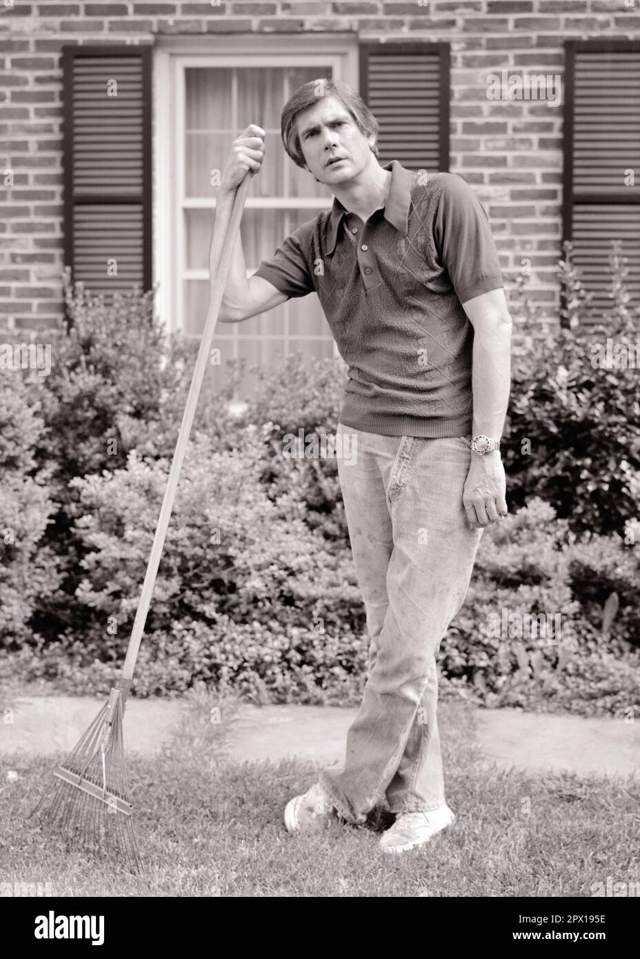 1970s MAN STANDING IN FRONT OF BRICK HOUSE LEANING ON RAKE TIRED LAZY ...