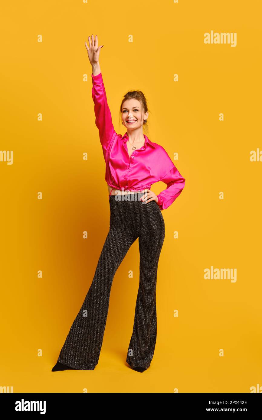 Joyful woman stands with her hand raised in a welcoming pose on yellow ...