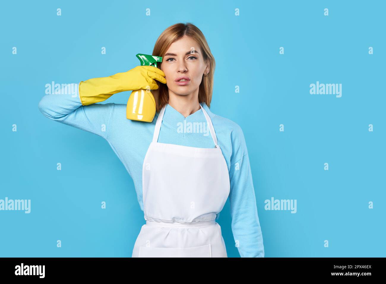 tired blonde woman in rubber gloves and cleaner apron holding detergent ...