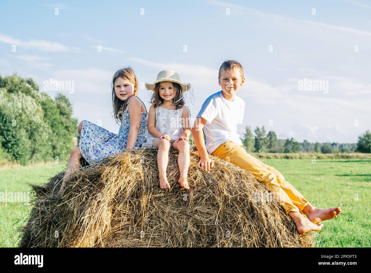 Portrait of three kids boy and two girls sitting on haystack in field ...