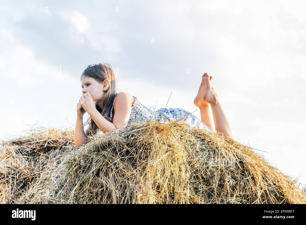 Thoughtful little girl lying on stomach at top of haystack. From below ...