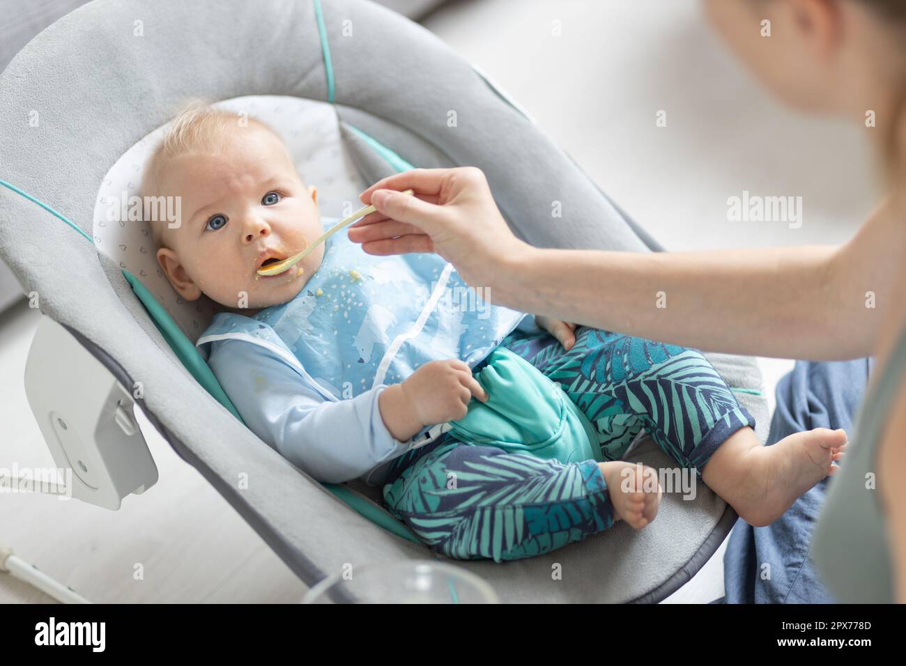 Mother spoon feeding her baby boy child in baby chair with fruit puree ...