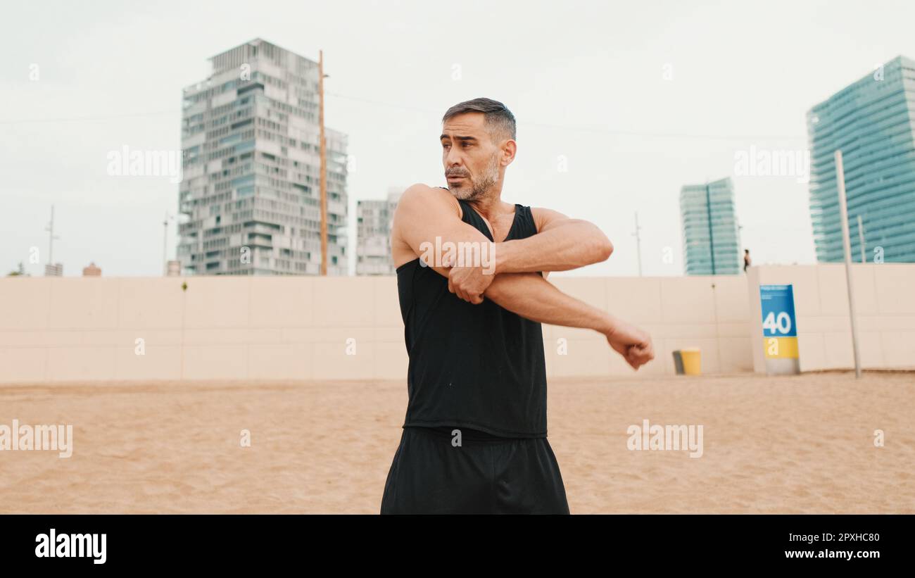 Athletic mature man stretches arm muscles while standing on the beach ...
