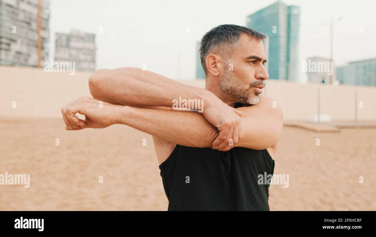 Athletic mature man stretches arm muscles while standing on the beach ...