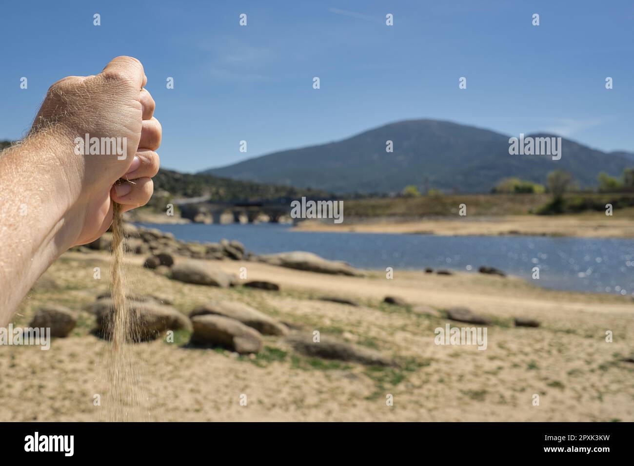 A man's hand pouring sand onto a shoreline in front of a tranquil lake ...