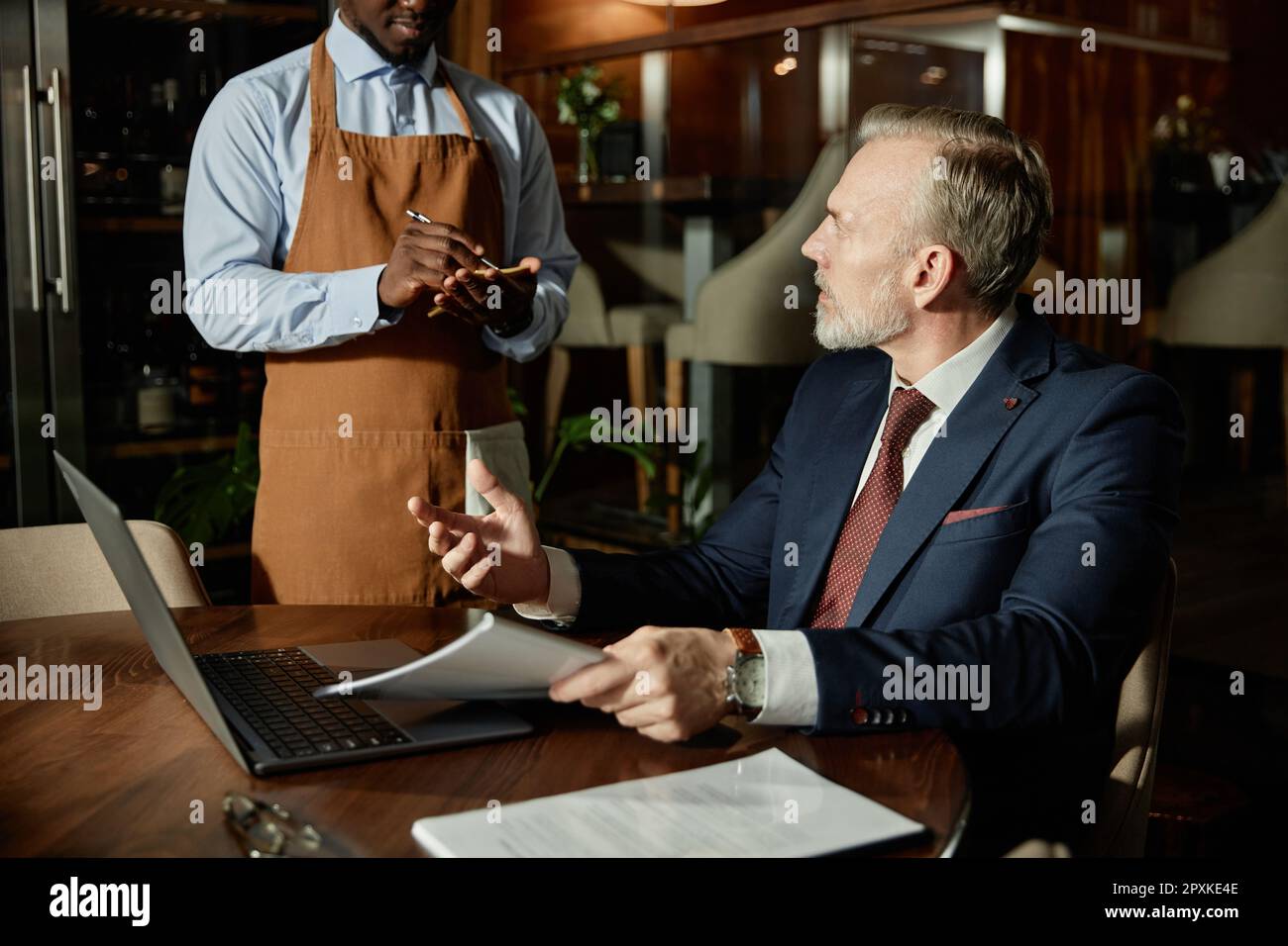 Mature businessman in suit making order to waiter while sitting at ...