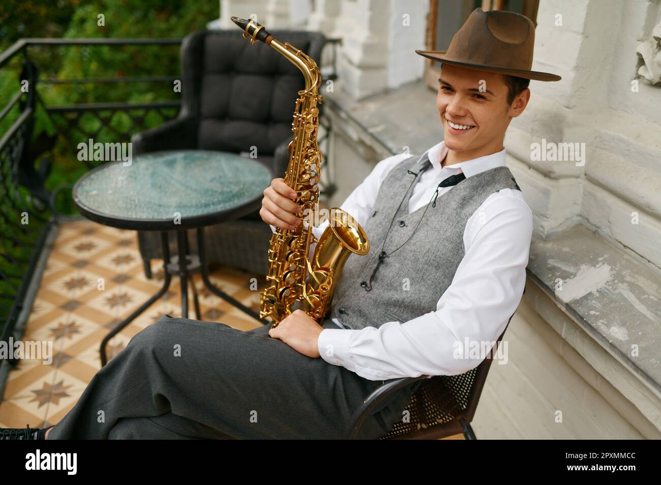 Smiling musician holding saxophone sitting on chair in balcony of his ...