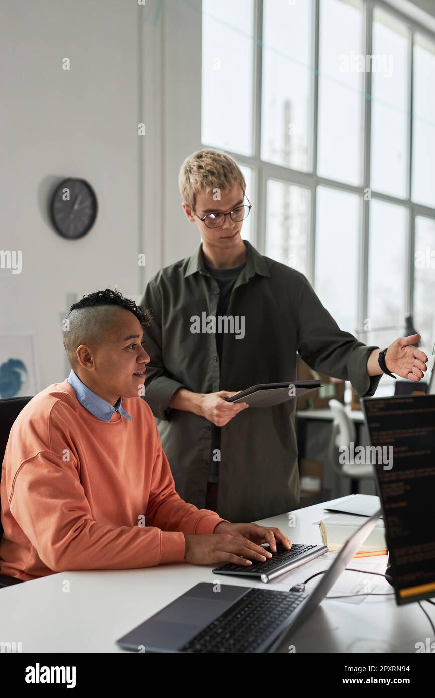 Vertical image of young colleagues discussing codes of software pointing at computer monitor ...