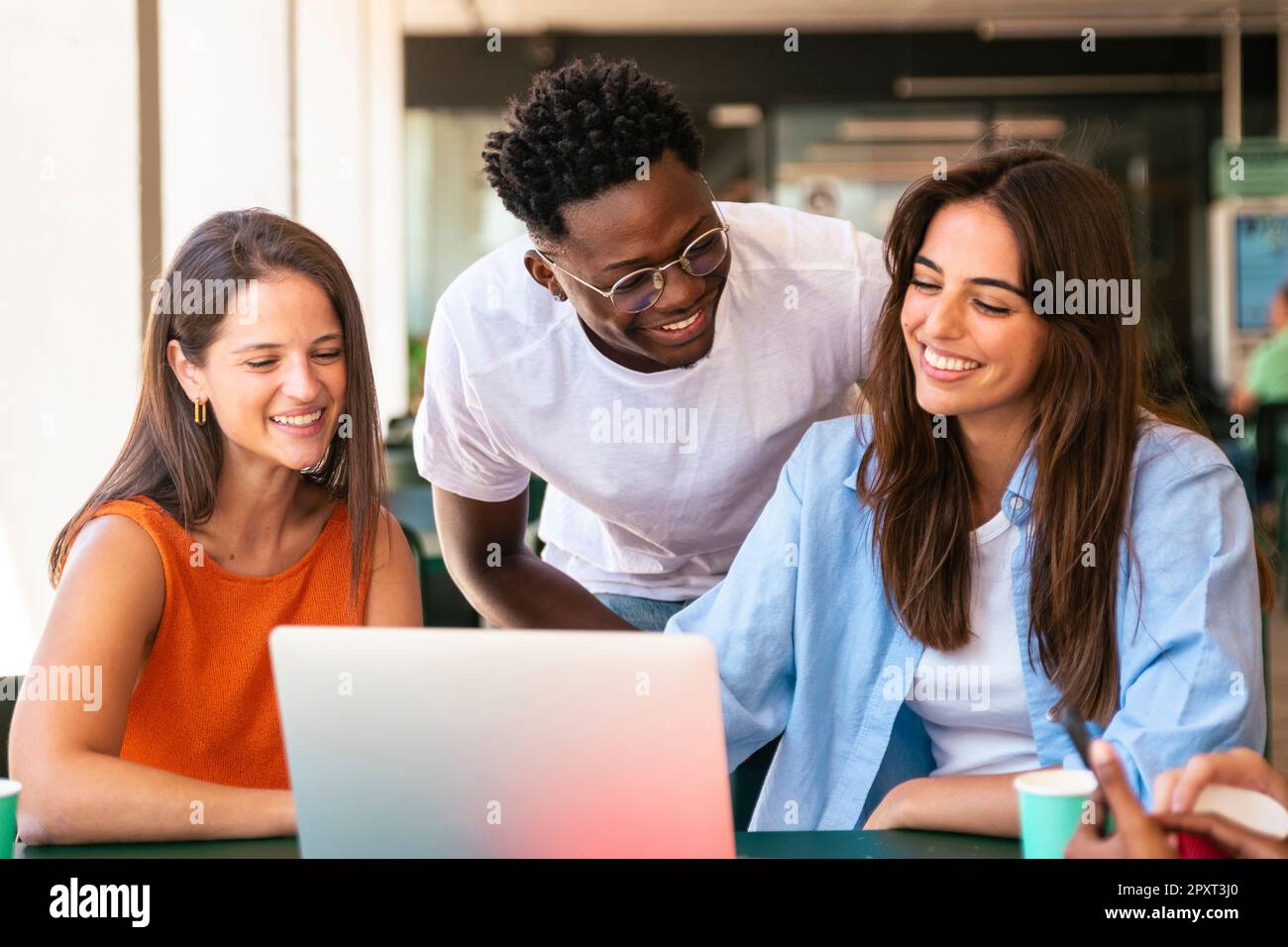 Group of happy multiracial students sitting in a cafe bar looking at laptop. Three cheerful ...