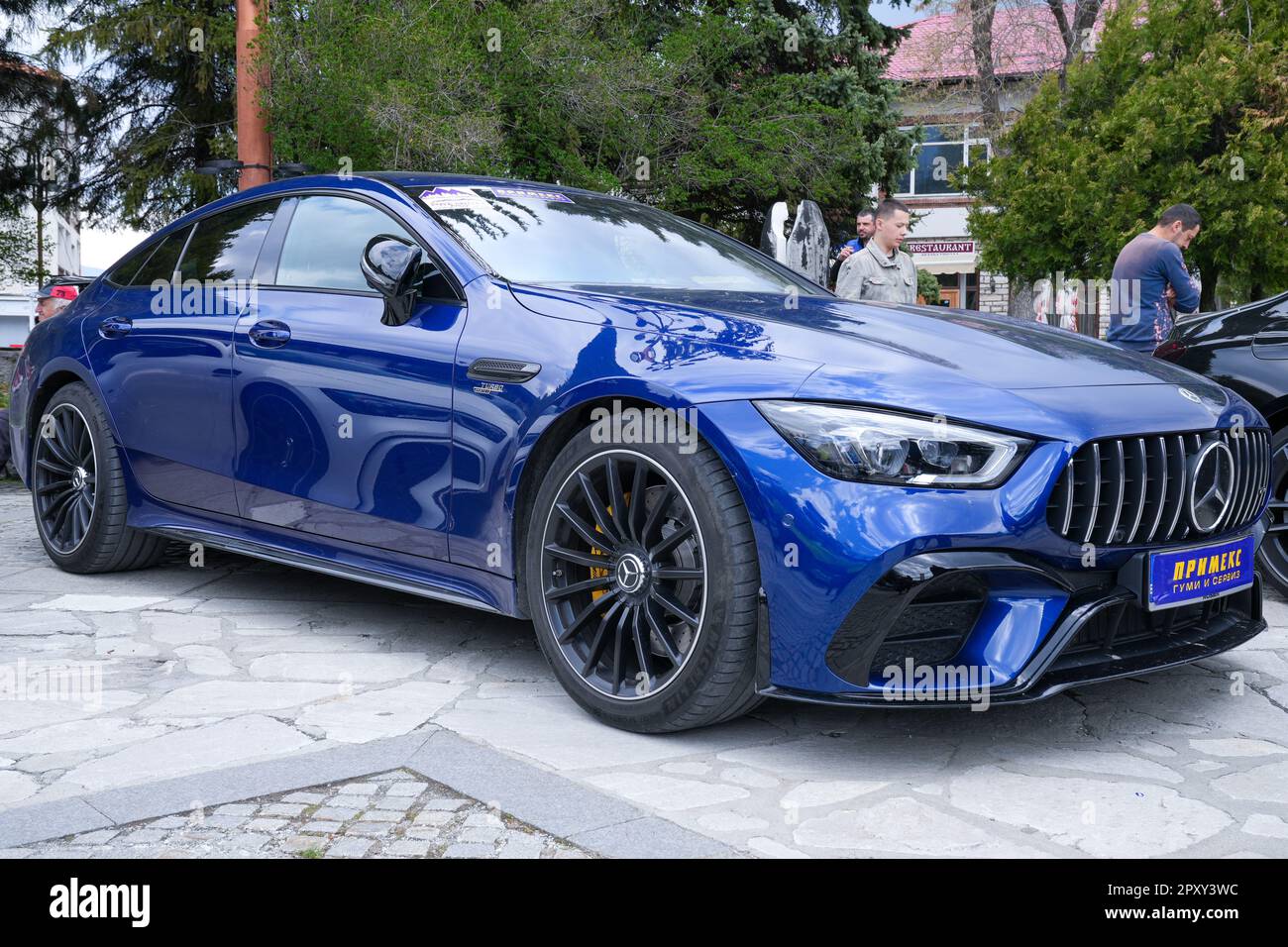 A blue Mercedes AMG GT43 sedan is parked in the foreground of an urban ...