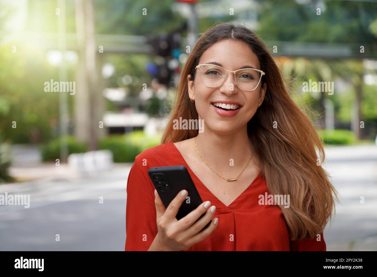 Brazilian woman looking at camera holding smartphone with blurred city ...