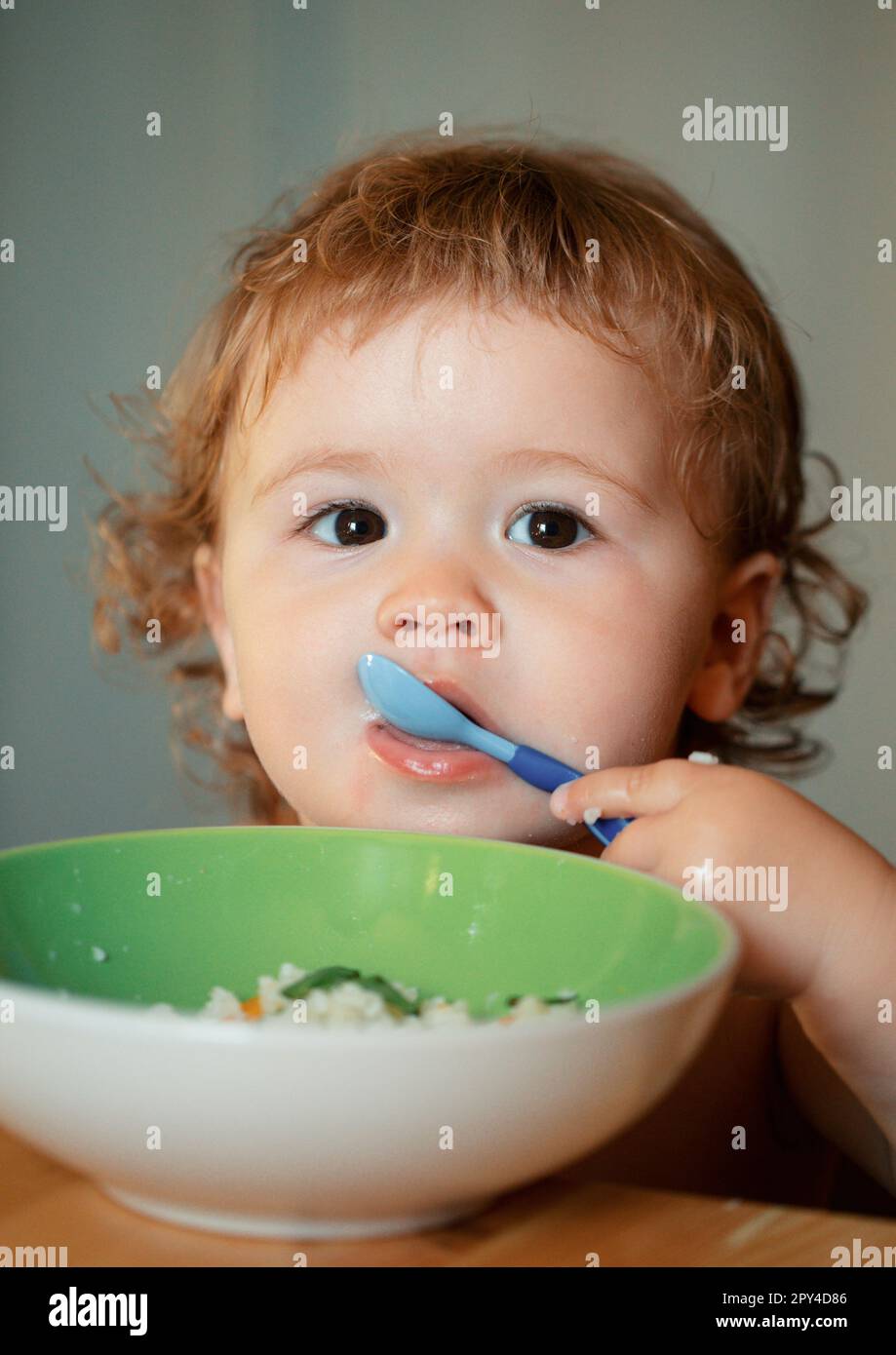 Funny baby eating food himself with a spoon on kitchen. Child nutrition ...