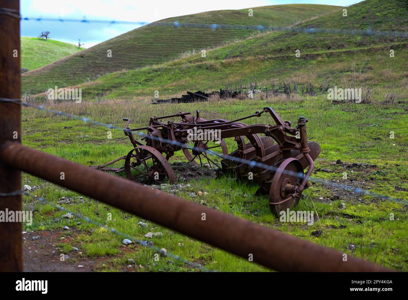 An old rusty plow sits behind a rusty metal fence with barb wire. The ...